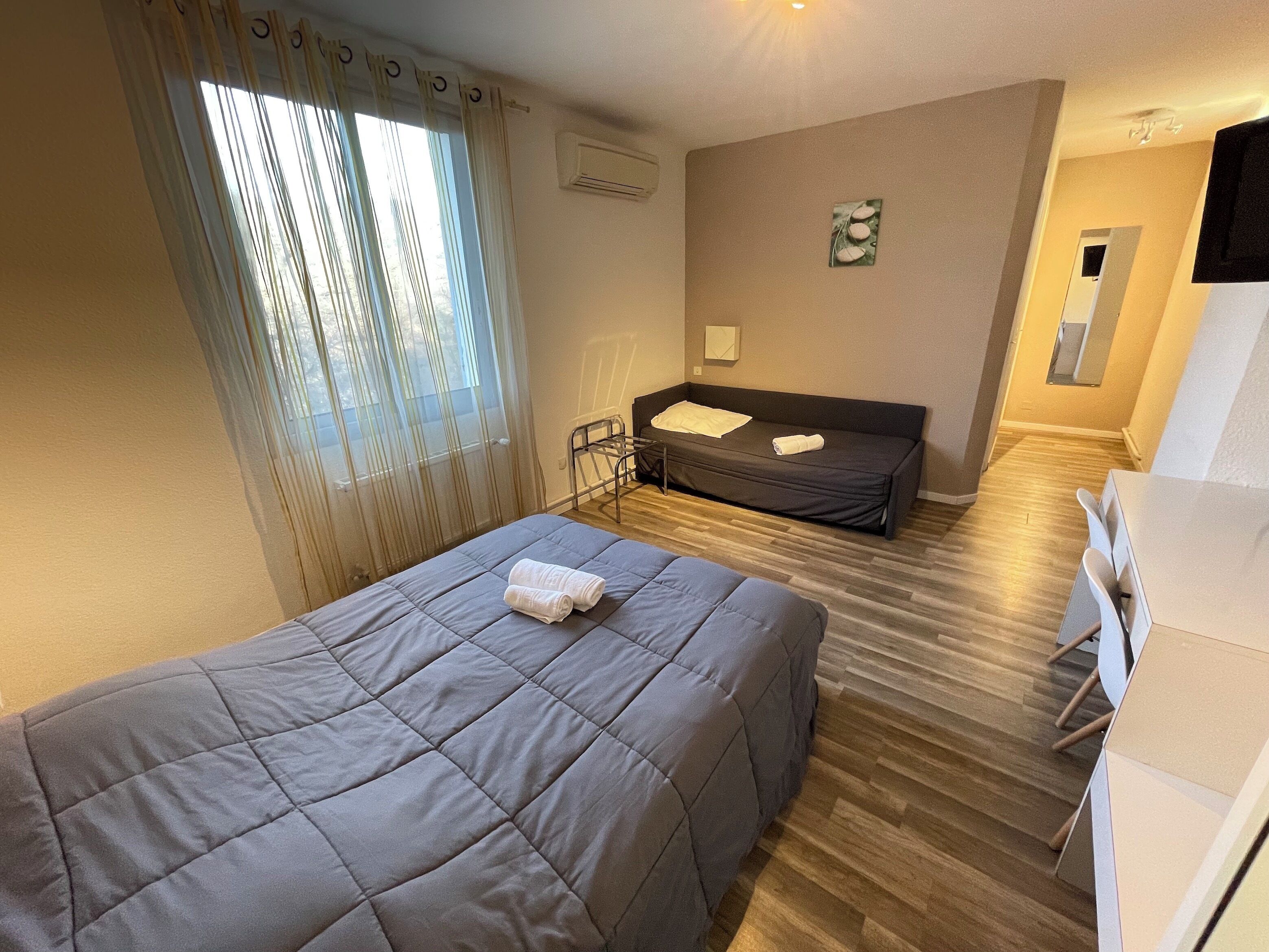 Quarto de hotel com cama, escrivaninha, cadeira, janela com cortinas, ar condicionado fixado na parede e uma pequena obra de arte.