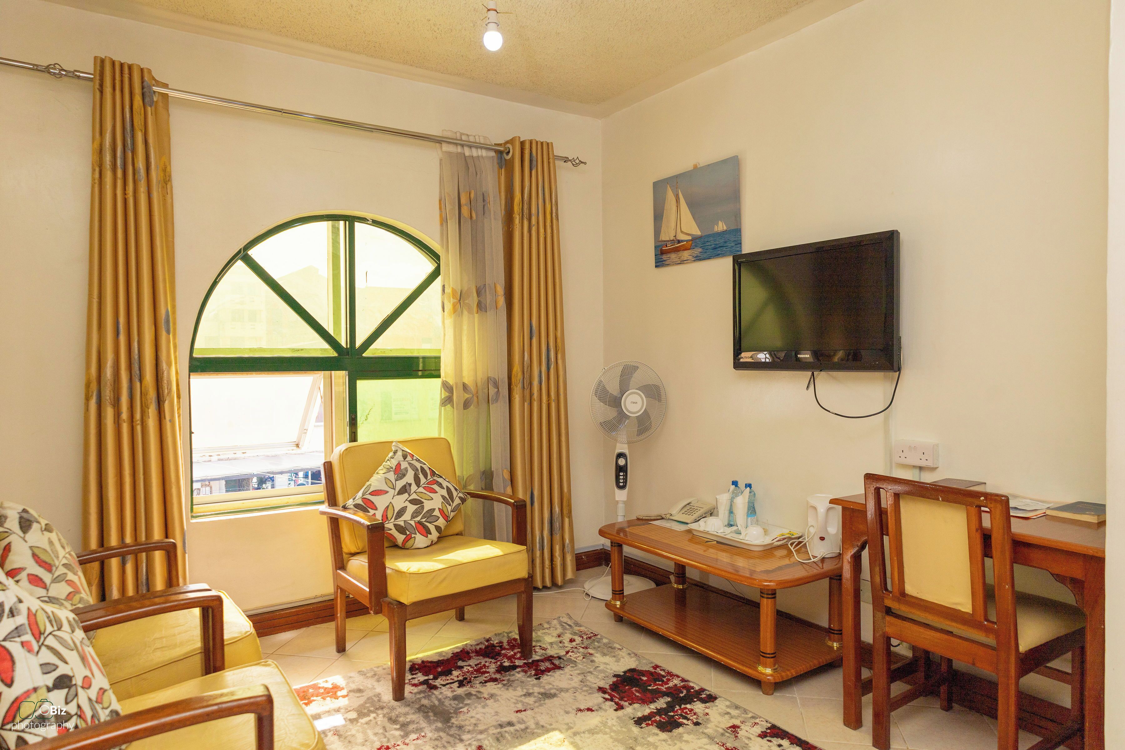 Quarto com janela grande, televisão, ventilador e escrivaninha com cadeira.