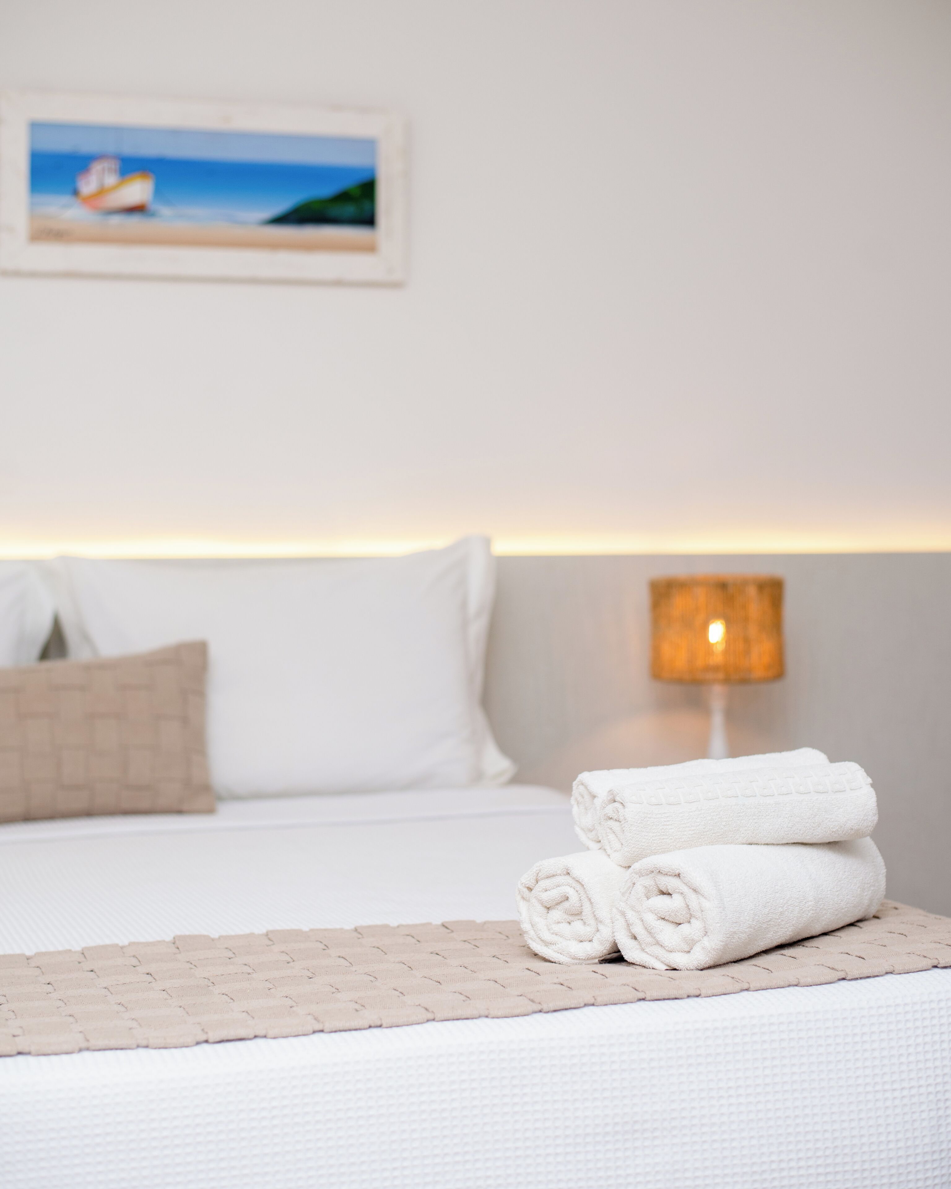 Cama bem arrumada com toalhas brancas dobradas, um travesseiro branco, um mantão bege e um quadro com uma imagem de barco na parede.