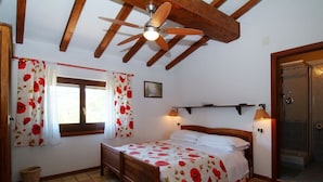Quarto com cama de madeira, ventoinha no teto, janela com cortinas florais, abajur e área de chuveiro.