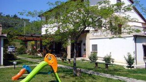 Uma área residencial com uma casa branca, um gramado verde e um parquinho infantil com escorrega e balanços.