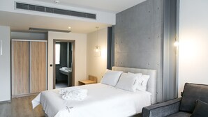 Quarto de hotel moderno com uma cama grande, um sofá cinzento, um guarda-roupa de madeira e uma pequena mesa de cabeceira.