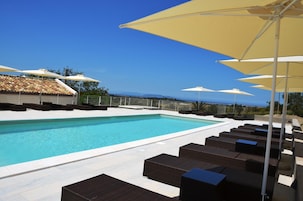 Uma piscina com espreguiçadeiras e guarda-sóis, um telhado telhado e vista para o mar.