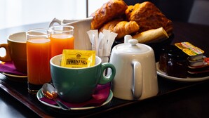 Bandeja de café da manhã com croissants, suco, chá Twinings e um bule sobre uma mesa, com um abajur ao fundo.