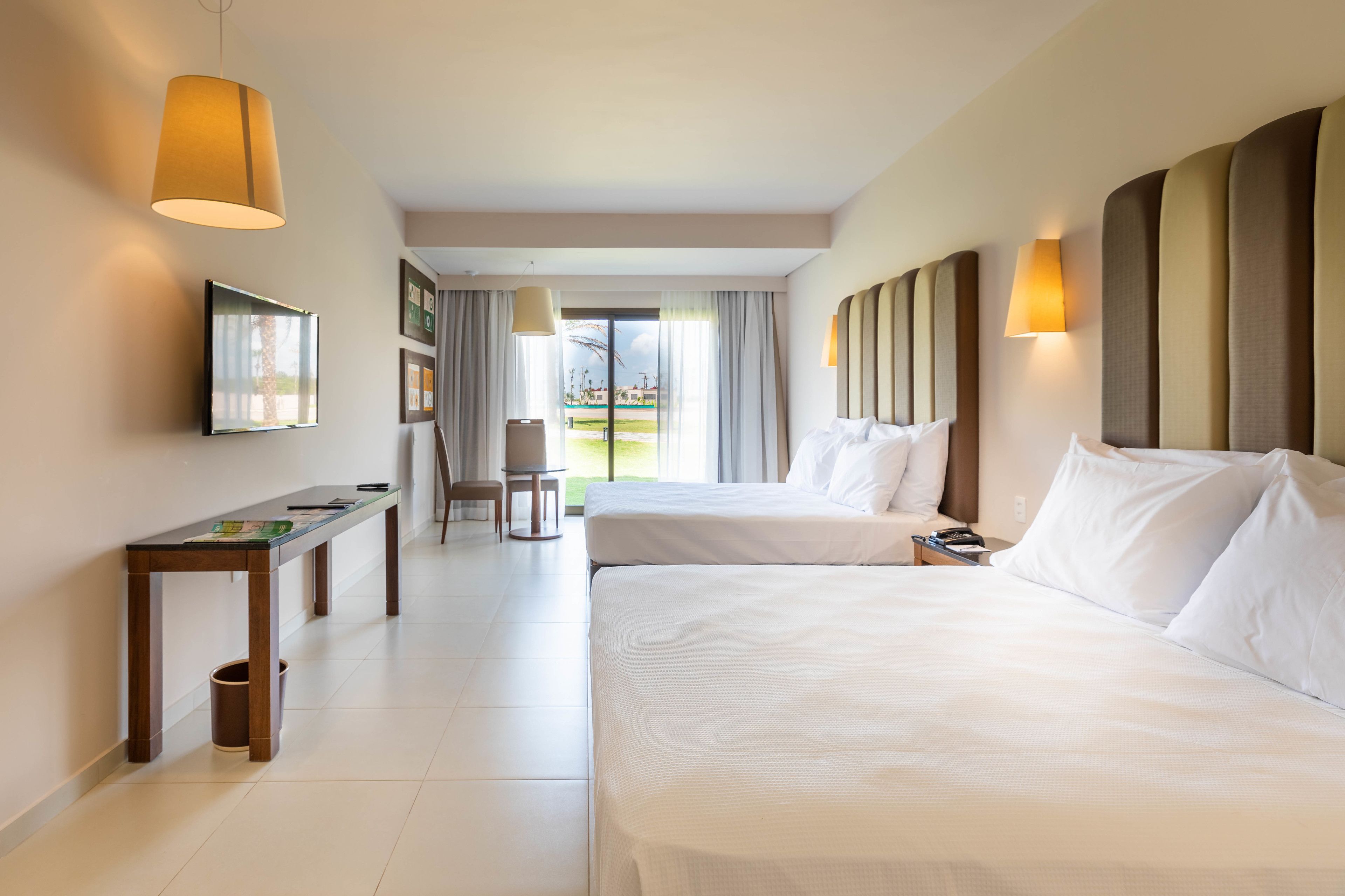 Quarto de hotel com duas camas, uma escrivaninha, uma cadeira e vista para o exterior.