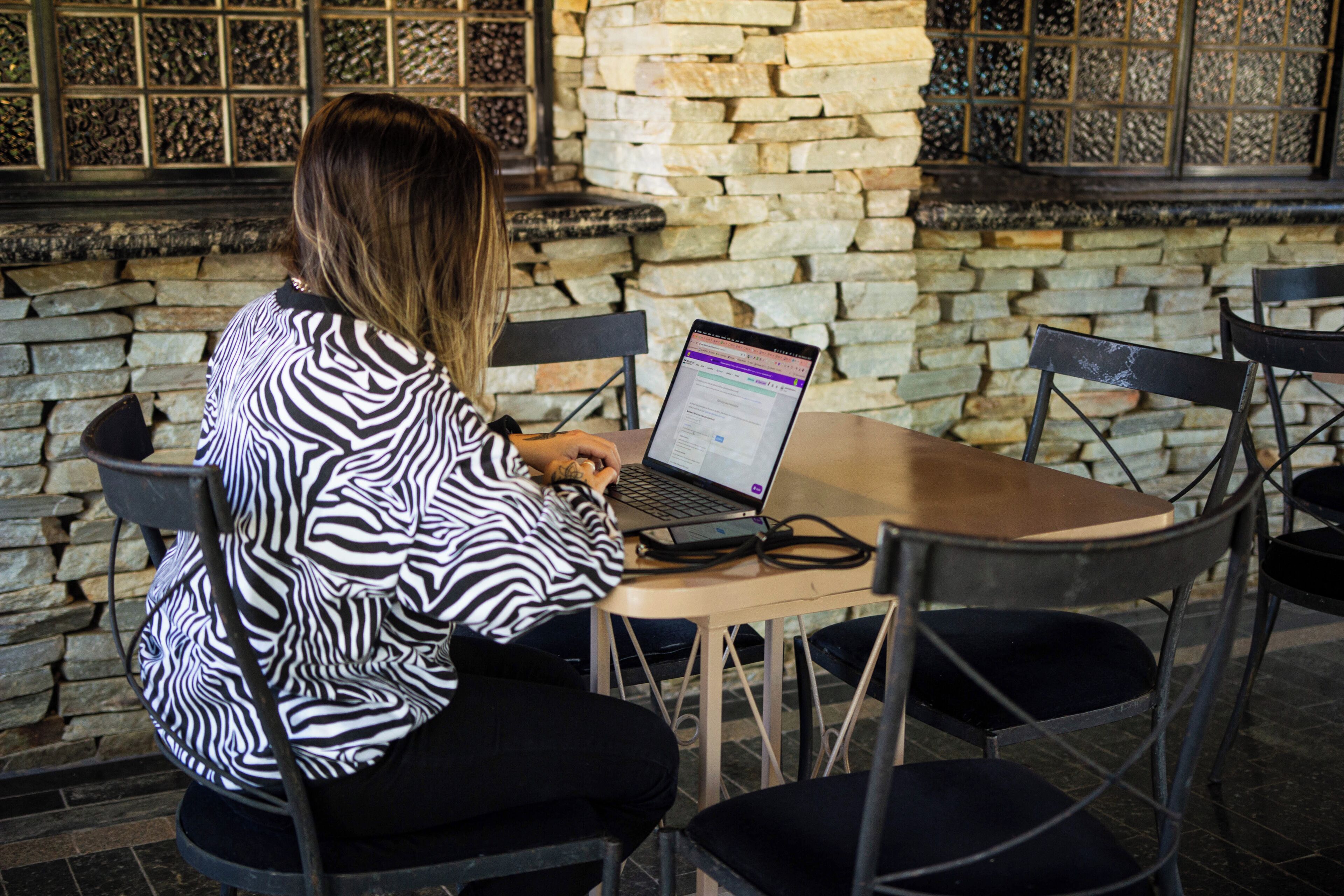 Uma pessoa sentada à mesa com um laptop, cercada por cadeiras, em um quarto com paredes de pedra e uma janela com padrão quadriculado.