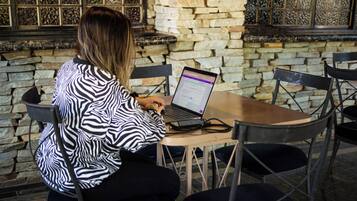 Uma pessoa sentada à mesa com um laptop, cercada por cadeiras, em um quarto com paredes de pedra e uma janela com padrão quadriculado.