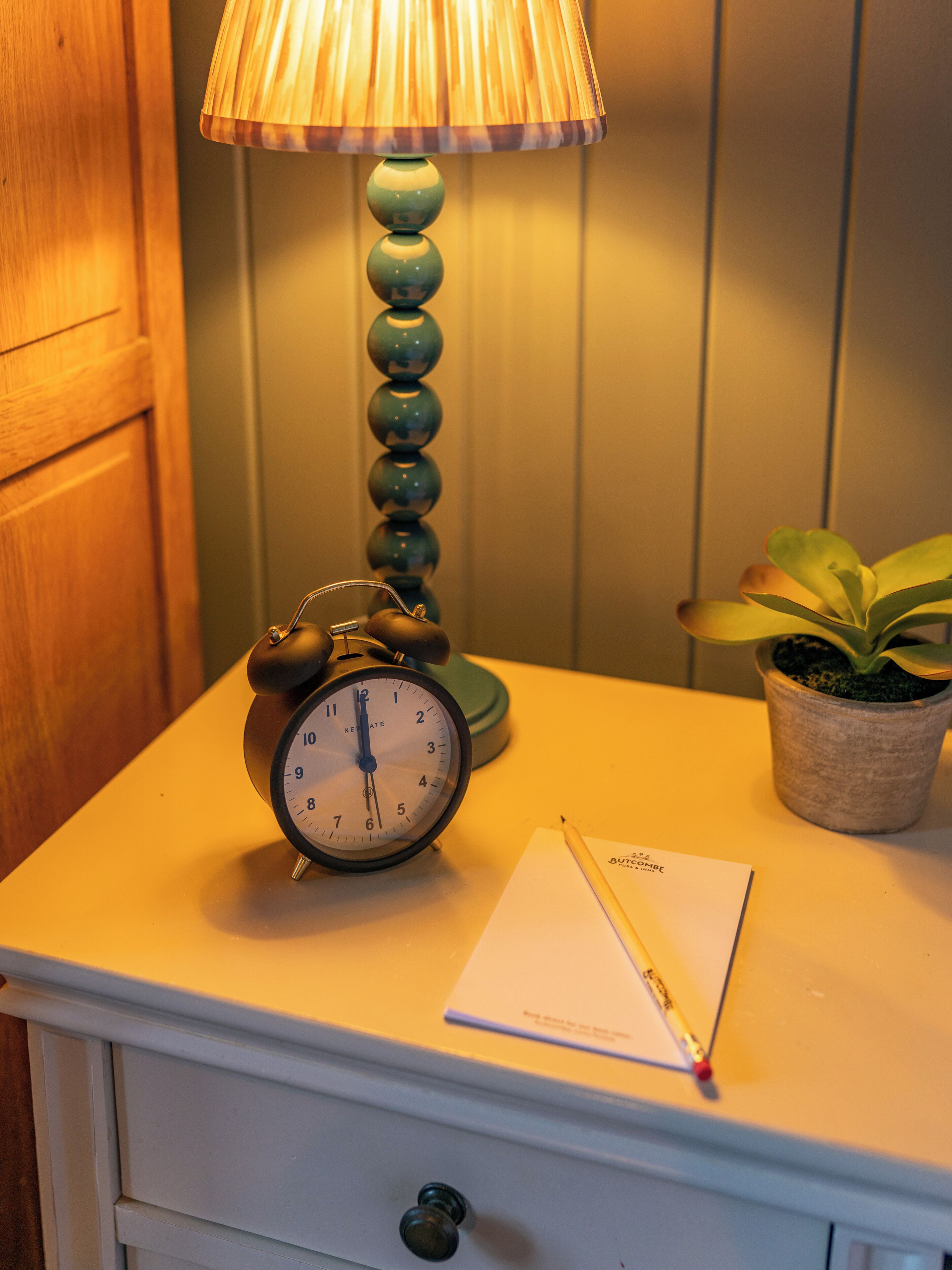 Uma mesa de cabeceira com uma luminária verde, um relógio, uma planta e um bloco de notas.