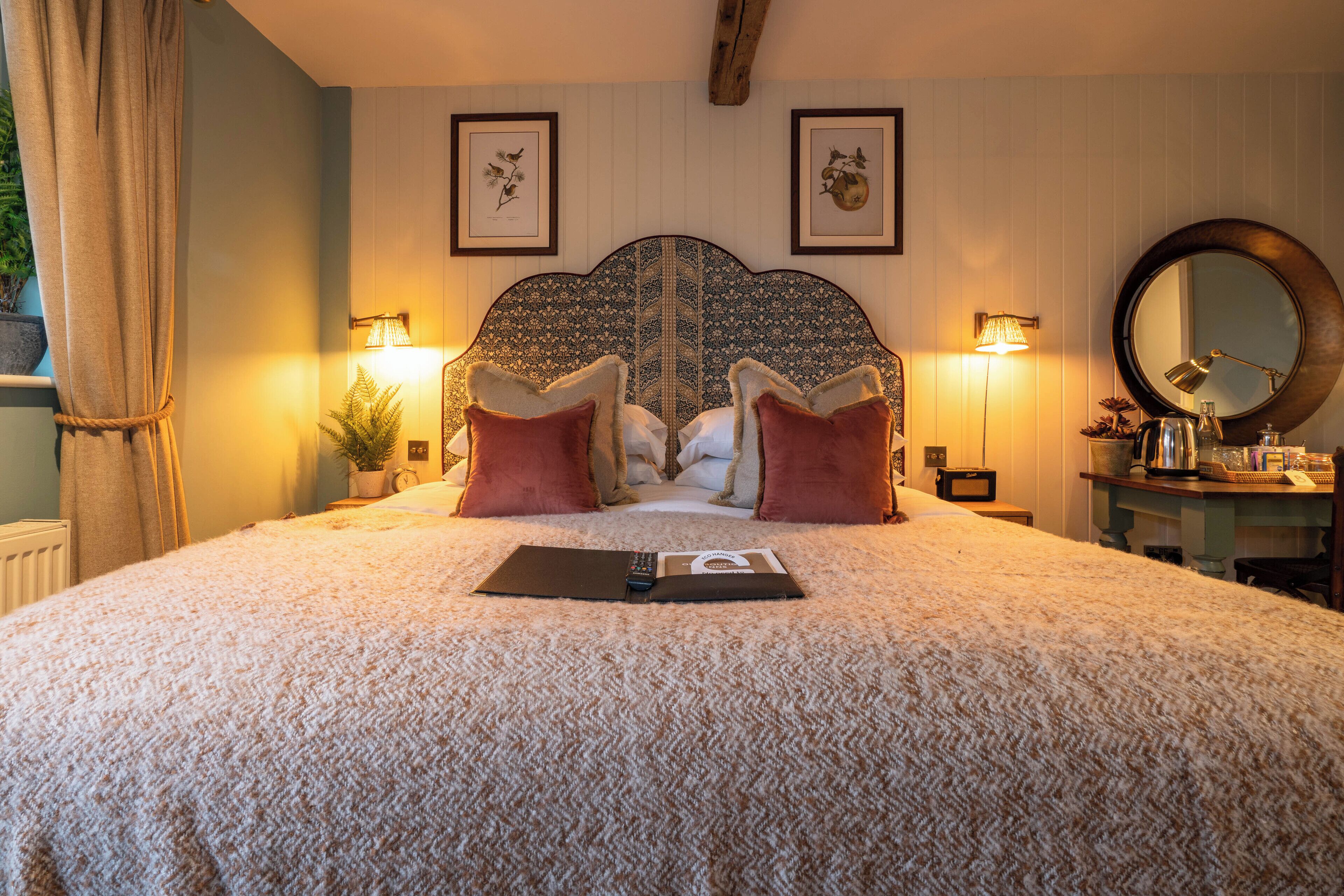 Cama bem arrumada com cabeceira, duas luminárias de apoio e mesa de cabeceira com abajur e espelho.