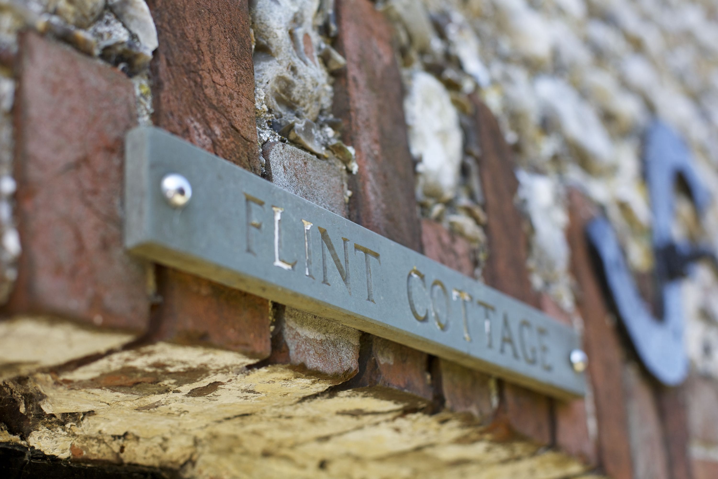 Placa de metal com a inscrição 'FLINT COTTAGE' fixada em uma parede de tijolos.