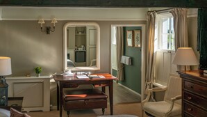 Quarto de hotel com cama, escrivaninha com cadeira, penteadeira, espelho e janela com cortinas.