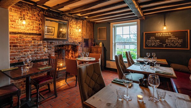Um restaurante acolhedor com paredes de tijolo, vigas de madeira e um quadro com o menu. As mesas estão montadas com copos e talheres.