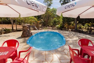 Piscina com azulejos de pedra, rodeada por cadeiras de plástico vermelhas e guarda-sóis.