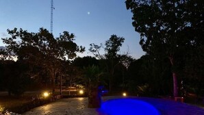 Área de piscina ao ar livre ao anoitecer, com piso de pedra e assentos de madeira.