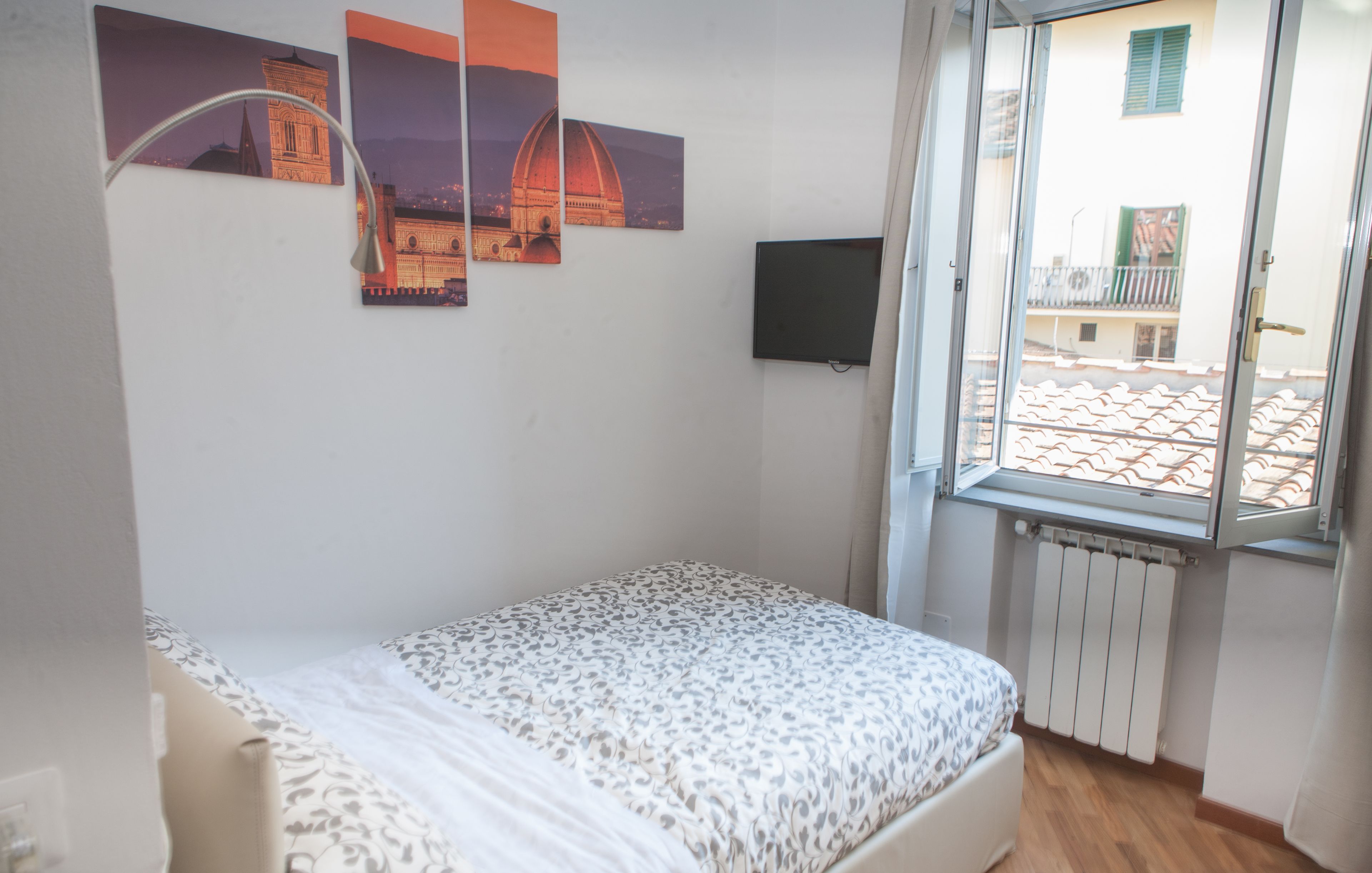 Quarto com cama, janela com vista para edifícios e três quadros pendurados na parede.