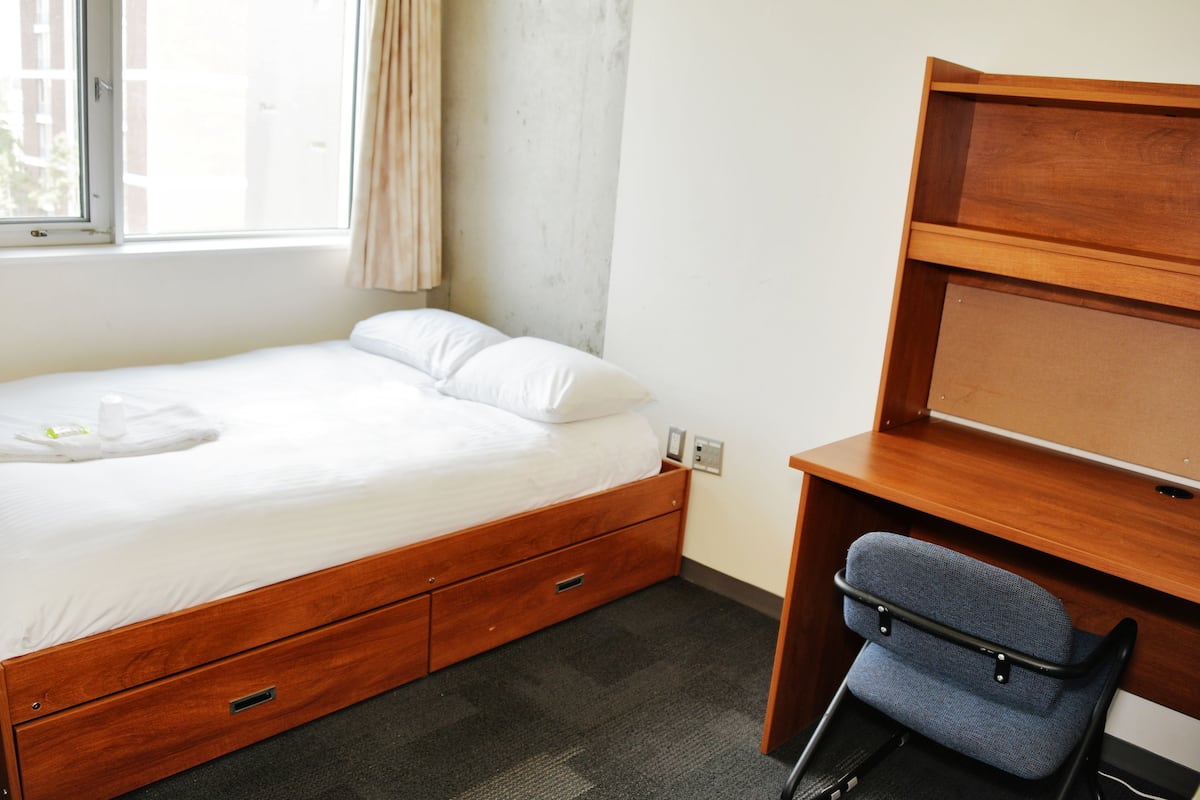 Uma cama de solteiro com estrutura de madeira, uma escrivaninha com cadeira e uma janela com cortinas.