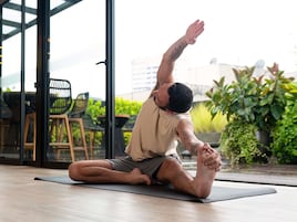 Uma pessoa fazendo uma postura de yoga em um tapete, em ambiente interno, com vista para área de estar externa e vegetação.