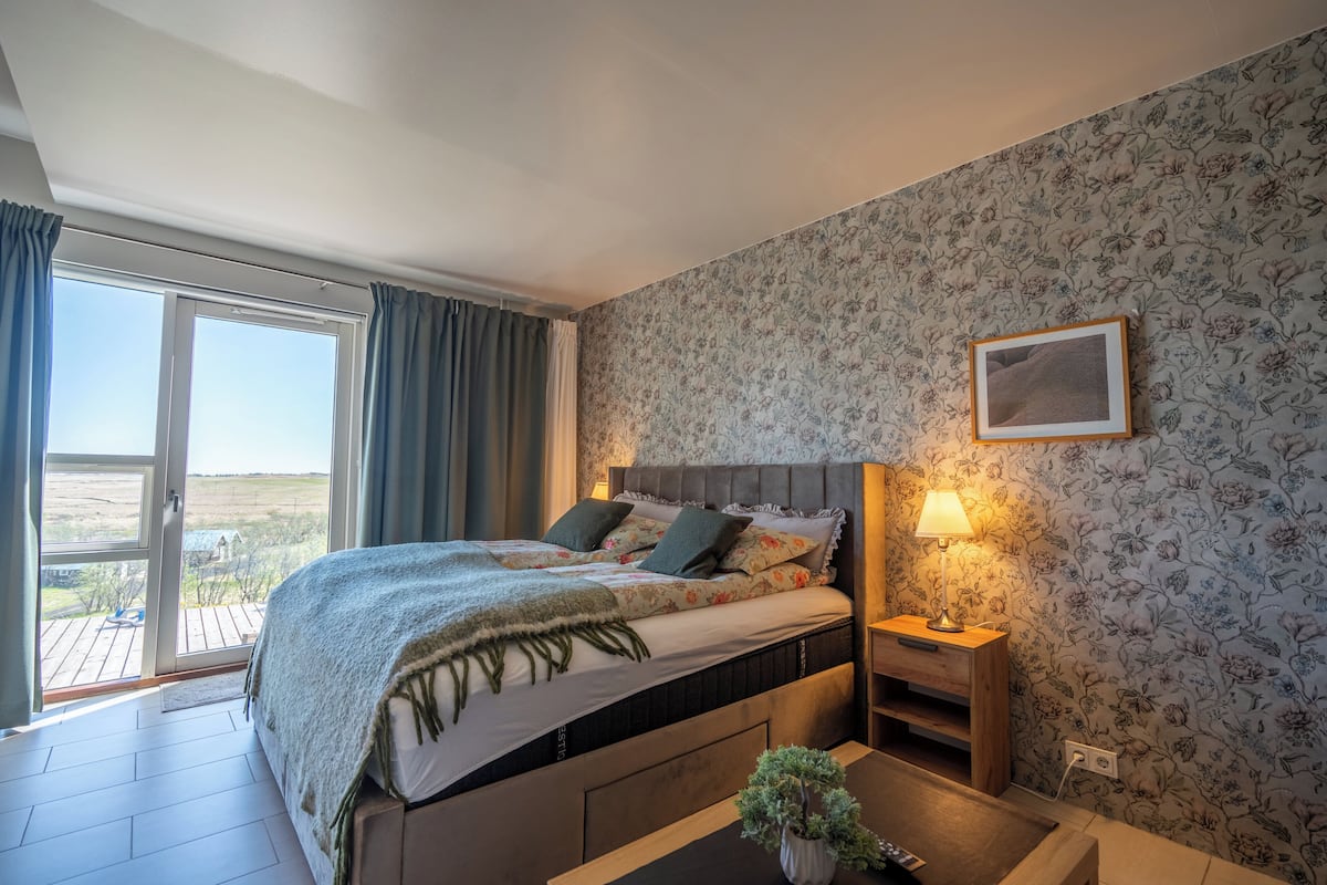 Quarto com cama grande, mesa de cabeceira com abajur, janela com cortinas e papel de parede floral.