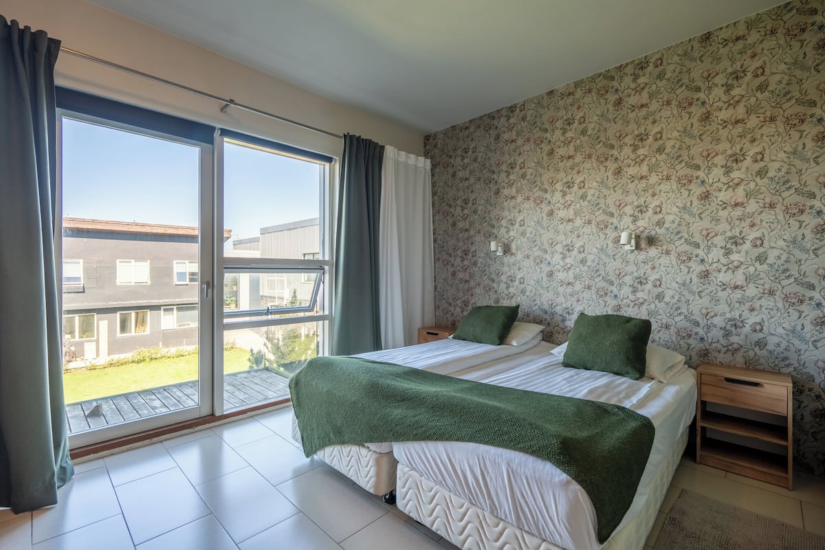 Quarto com uma grande janela, cama com roupa de cama verde, mesa de cabeceira e papel de parede floral.