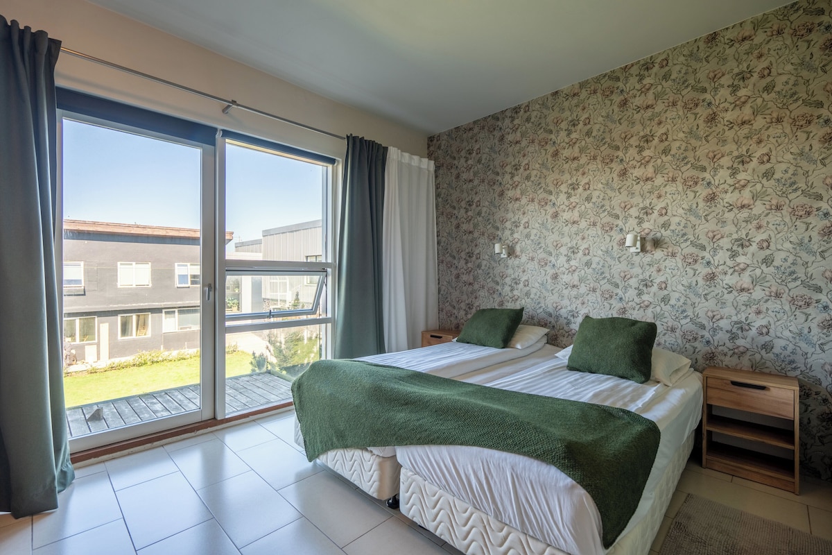 Quarto com uma grande janela, cama com roupa de cama verde, mesa de cabeceira e papel de parede floral.