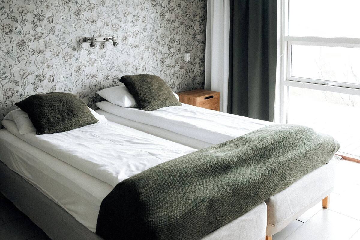 Uma cama de casal com um cobertor verde, uma mesa de cabeceira com abajur e uma janela com cortinas.