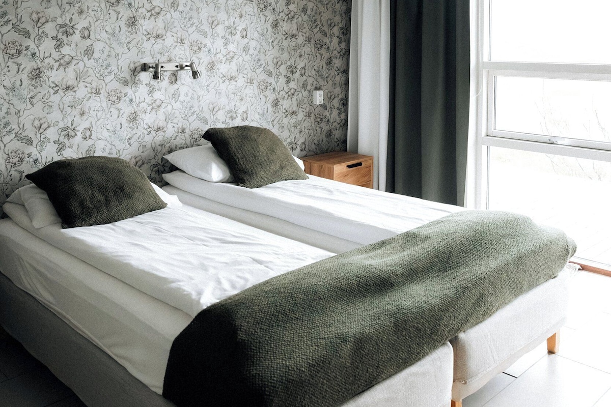 Uma cama de casal com um cobertor verde, uma mesa de cabeceira com abajur e uma janela com cortinas.
