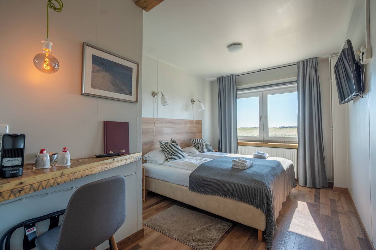 Quarto de hotel com cama, escrivaninha com cafeteira, cadeira, janela com cortinas e um quadro na parede.