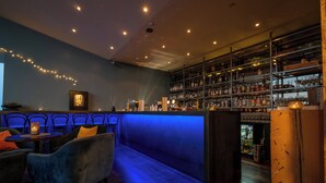 Uma área de bar com balcão iluminado em azul, assentos com almofadas e prateleiras cheias de garrafas.