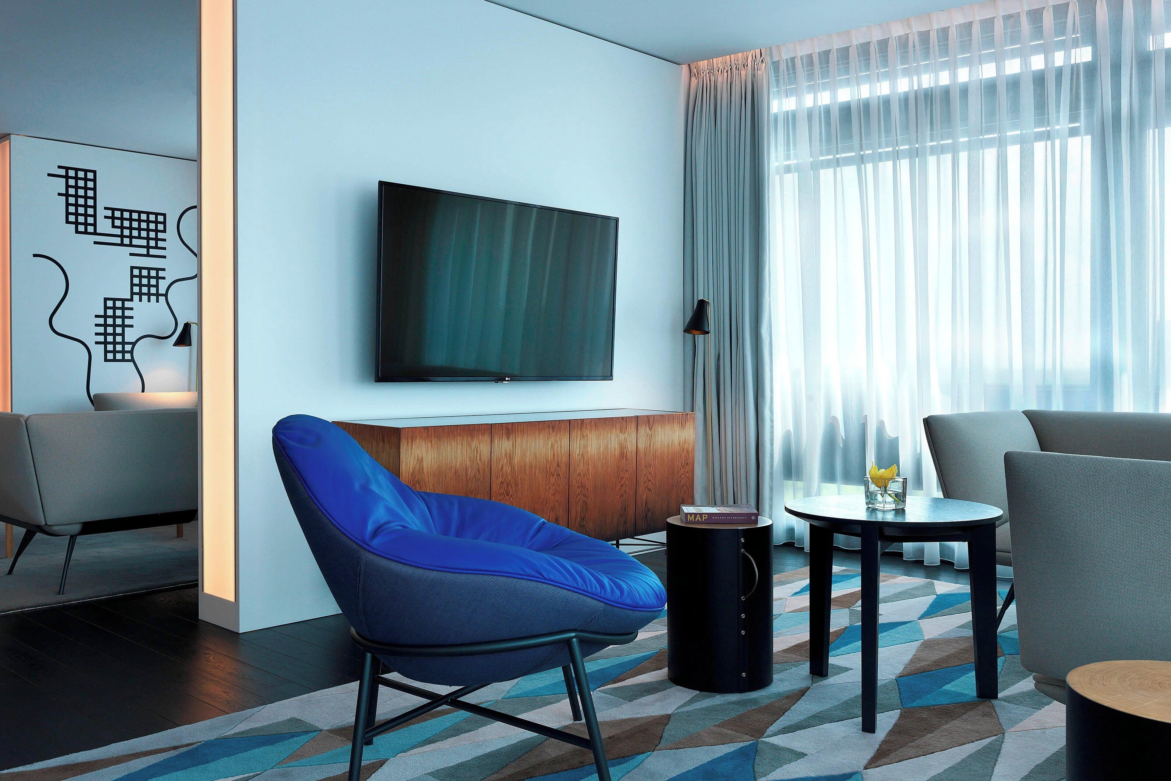 Quarto de hotel moderno com uma grande TV de tela plana na parede, uma poltrona de couro azul, uma pequena mesa redonda com abajur e um tapete estampado.