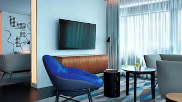 Quarto de hotel moderno com uma grande TV de tela plana na parede, uma poltrona de couro azul, uma pequena mesa redonda com abajur e um tapete estampado.