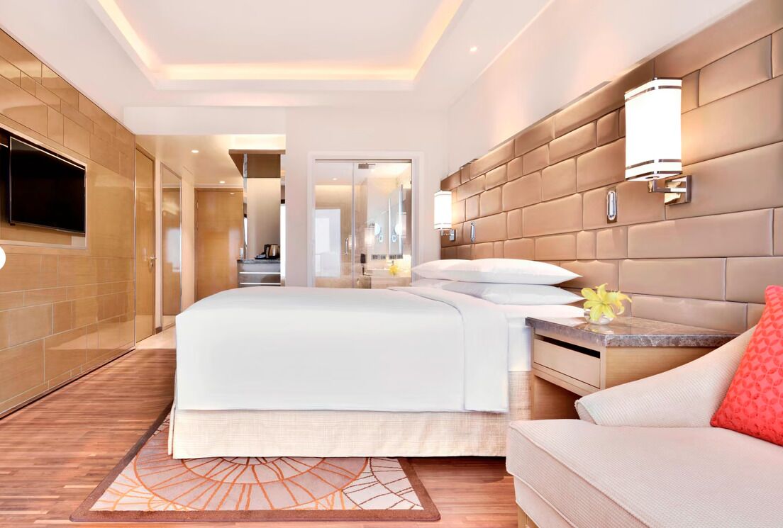 Quarto de hotel moderno com uma cama grande, área de estar, televisão de tela plana e banheiro visível através de uma porta de vidro.