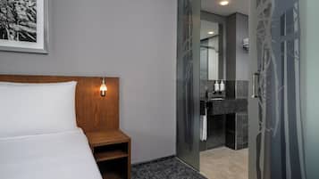 Quarto de hotel moderno com cabeceira de madeira, mesa de cabeceira, criadeira, uma cama grande, luminária de parede, banheiro com porta de vidro e um espelho.