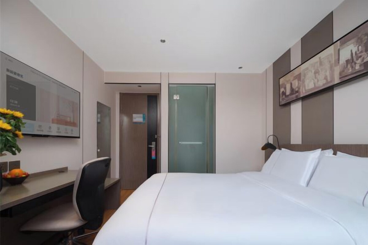 Quarto de hotel com cama, escrivaninha, cadeira e televisão presa à parede.