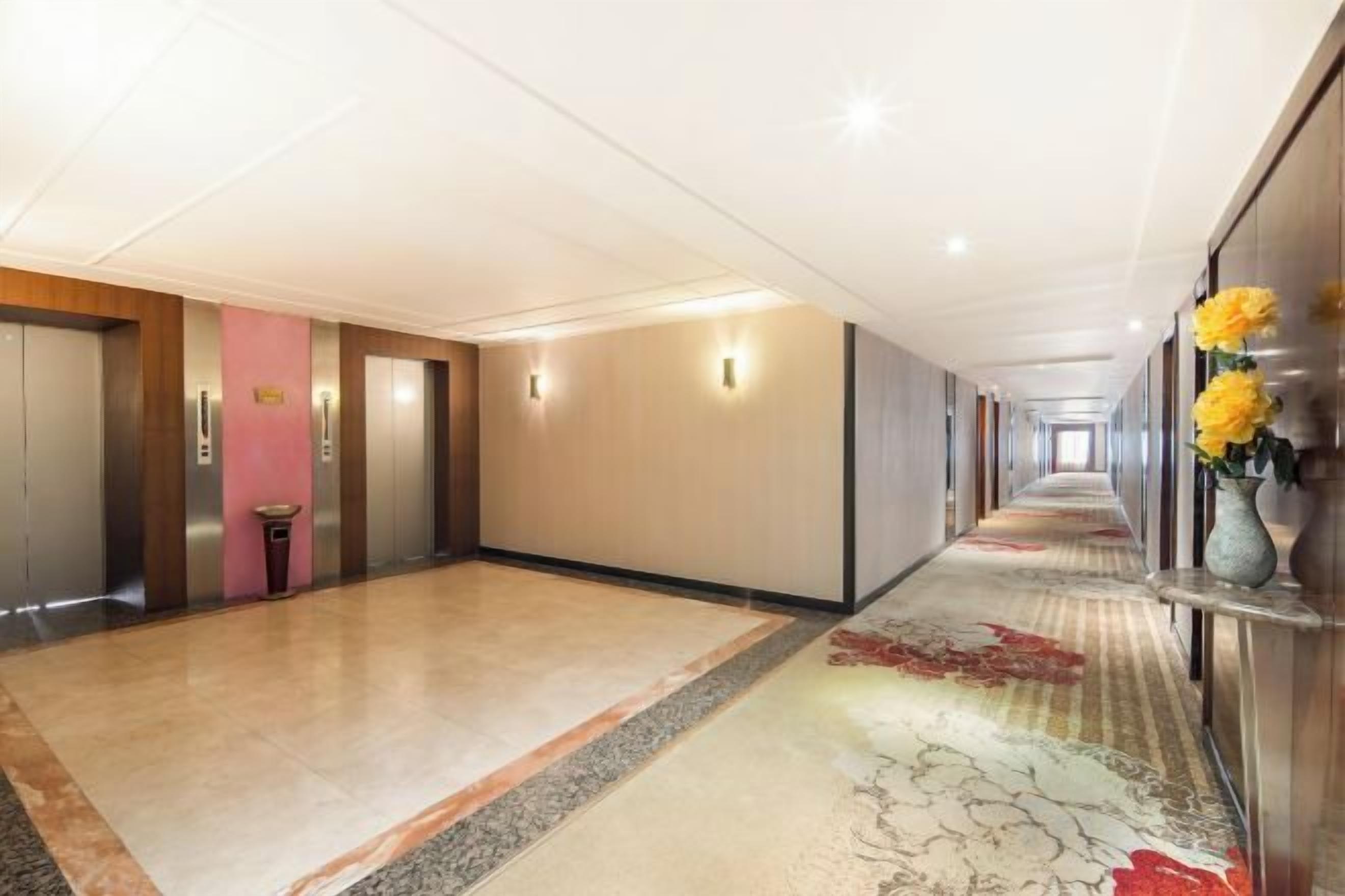 Corredor de hotel com piso carpetado, balcão de recepção e um vaso de flores fixado na parede.