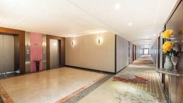 Corredor de hotel com piso carpetado, balcão de recepção e um vaso de flores fixado na parede.