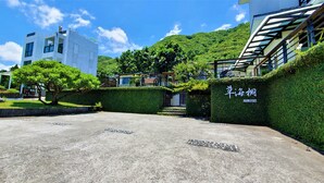 Um edifício moderno com telhado plano e grandes janelas, rodeado por vegetação exuberante e um espaço pavimentado com um caminho de pedra.