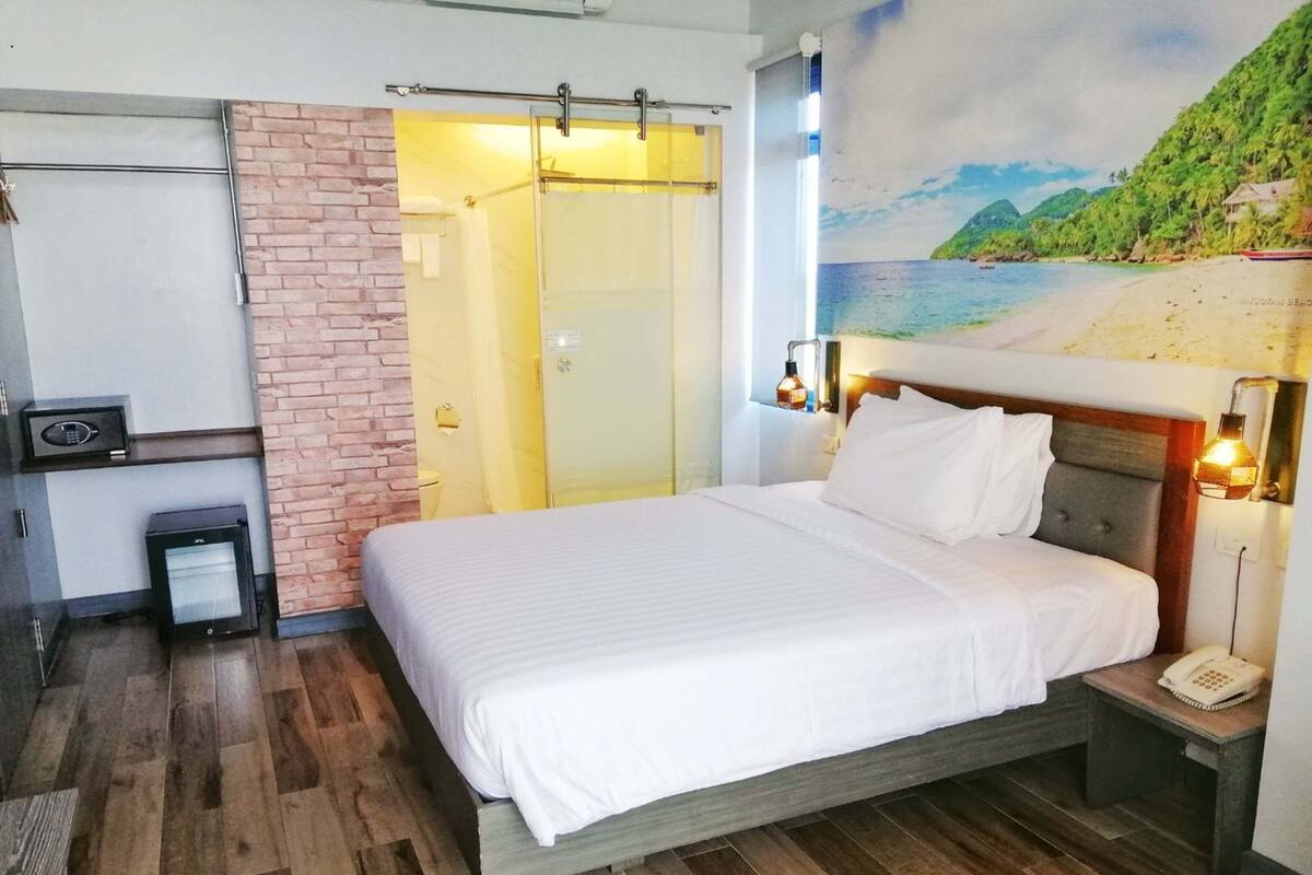 Quarto de hotel com uma cama, uma parede de tijolos, um banheiro, um quadro de uma praia e um telefone na mesa de cabeceira.