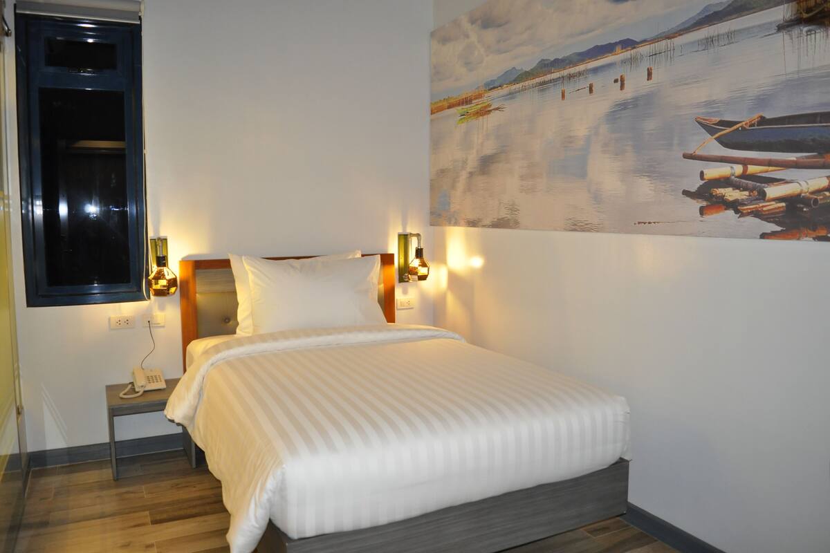 Quarto de hotel com cama, abajures, mesa de cabeceira com telefone e pintura de barcos na parede.