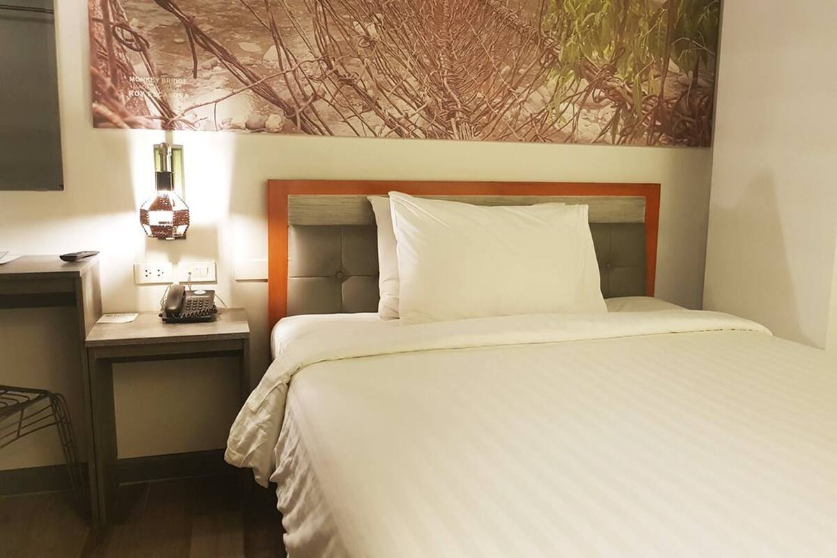 Quarto de hotel com cama, mesa de cabeceira e telefone, e luminária fixada na parede.