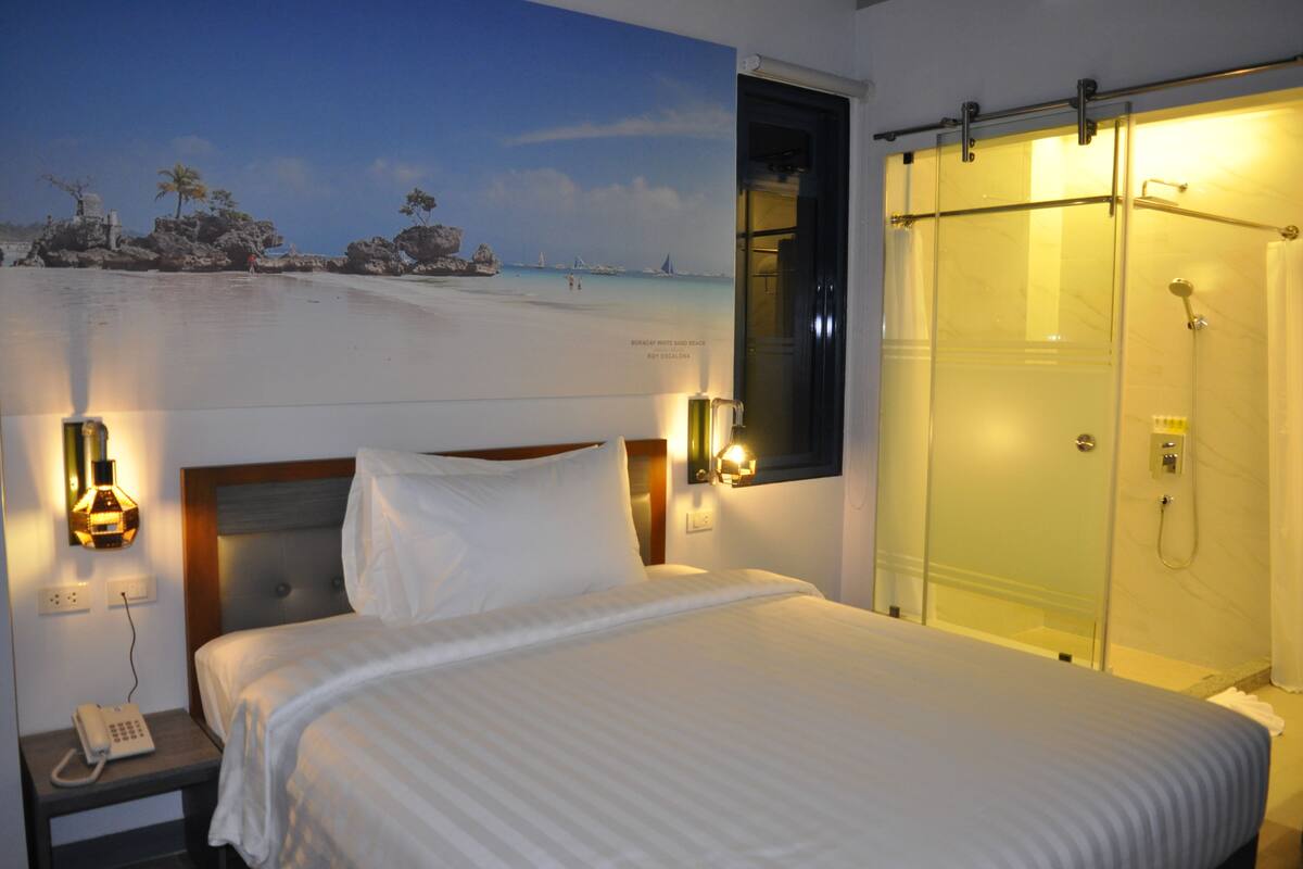 Quarto de hotel com uma cama, uma mesa de cabeceira com um telefone, um chuveiro com divisória de vidro e um mural de praia na parede.