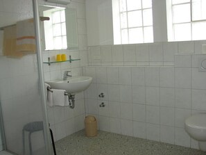 Casa de banho com lavatório branco, espelho e sanita. Dispõe de cabine de duche em vidro e um pequeno contentor de lixo.