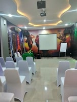 Uma sala de conferências com cadeiras brancas, uma tela de projetor e um palco com uma mesa verde.