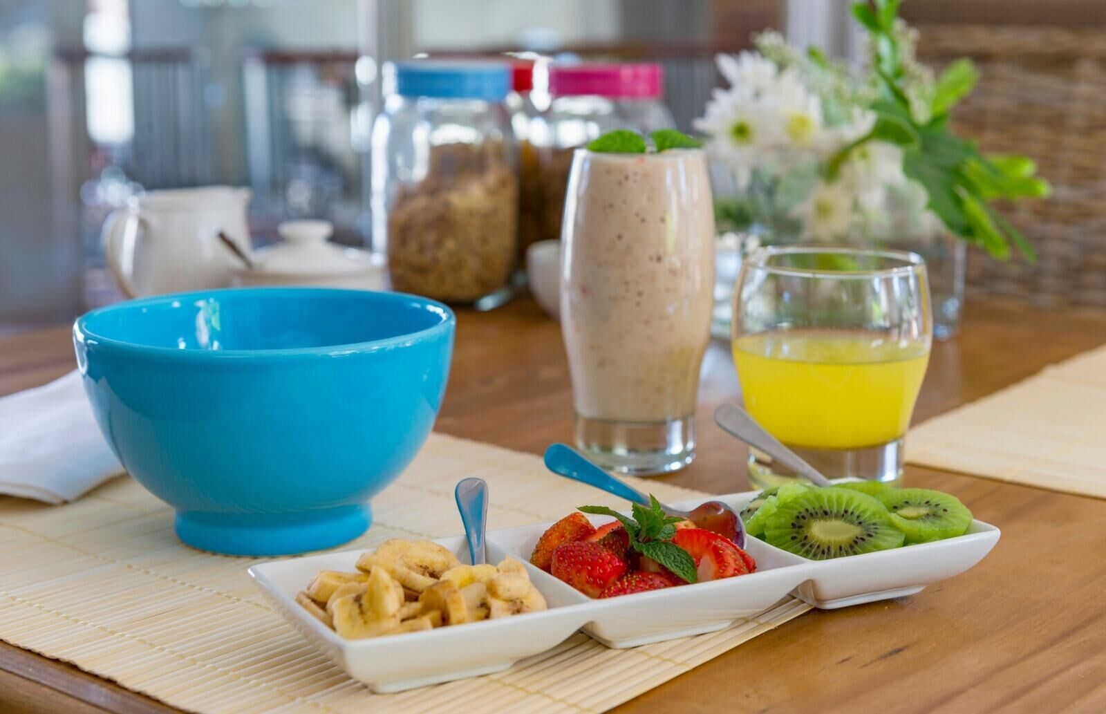 Uma mesa de café da manhã com uma tigela azul, um copo de suco de laranja e um prato de frutas, incluindo morangos e kiwi.