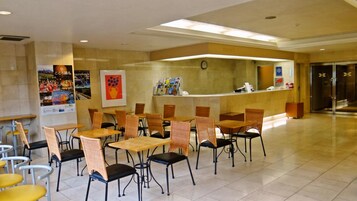 Um saguão de hotel com balcão de recepção, área de estar com mesas e cadeiras, e piso revestido de azulejos.