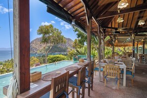 Uma área de refeições ao ar livre com mesa de madeira e vista para a piscina e oceano.