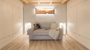 Uma sala de estar moderna com um sofá cinzento, duas mesinhas laterais, uma obra de arte com paisagem urbana emoldurada e teto de madeira.
