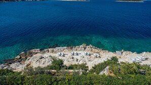 Vista costeira com mar azul claro, terreno rochoso e cadeiras de praia brancas.