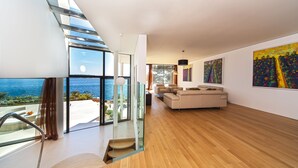Uma sala de estar ampla com uma grande janela com vista para o mar, uma escada que leva a um andar superior e um design de interiores moderno.