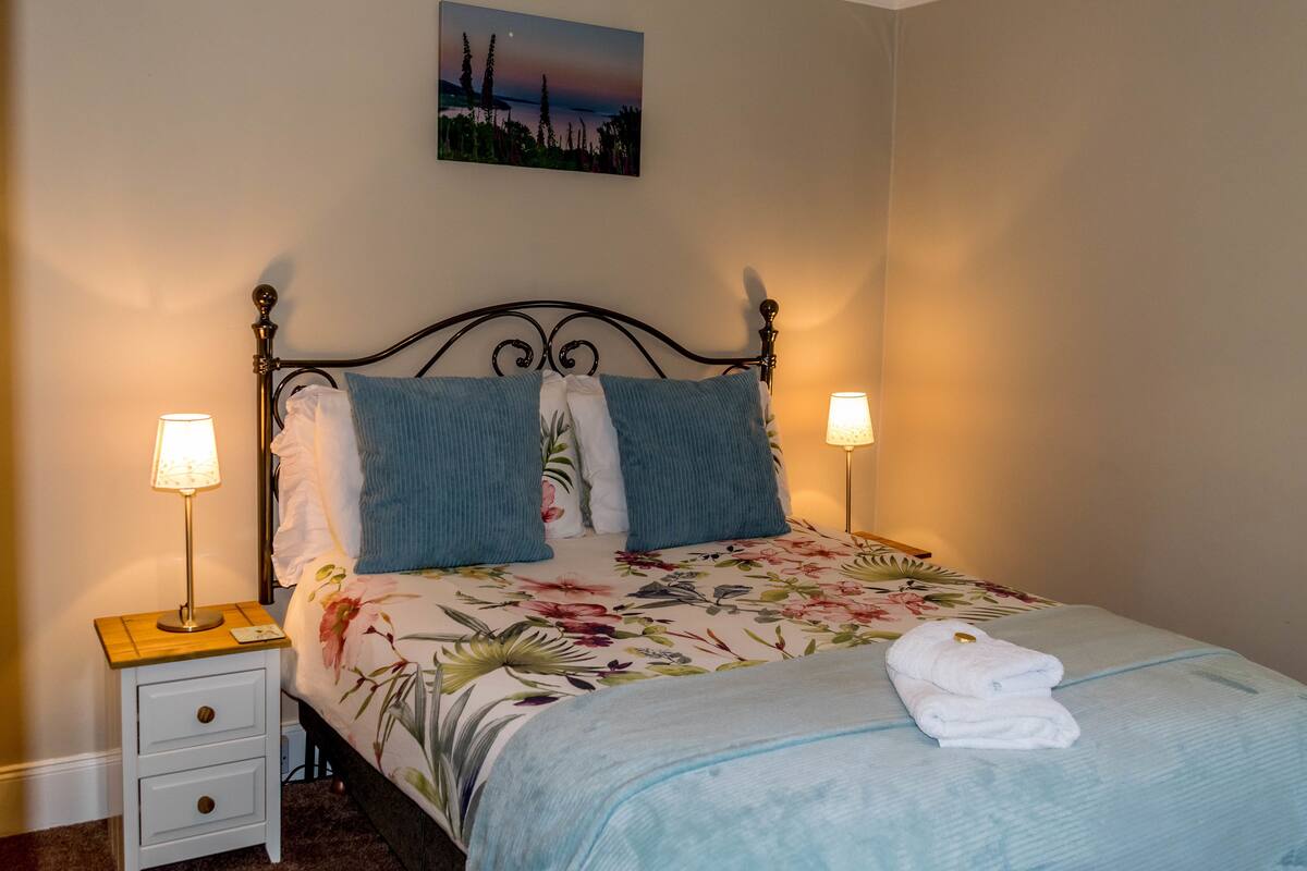 Cama bem arrumada com roupa de cama floral, duas luminárias de cabeceira e mesa de cabeceira com abajur.