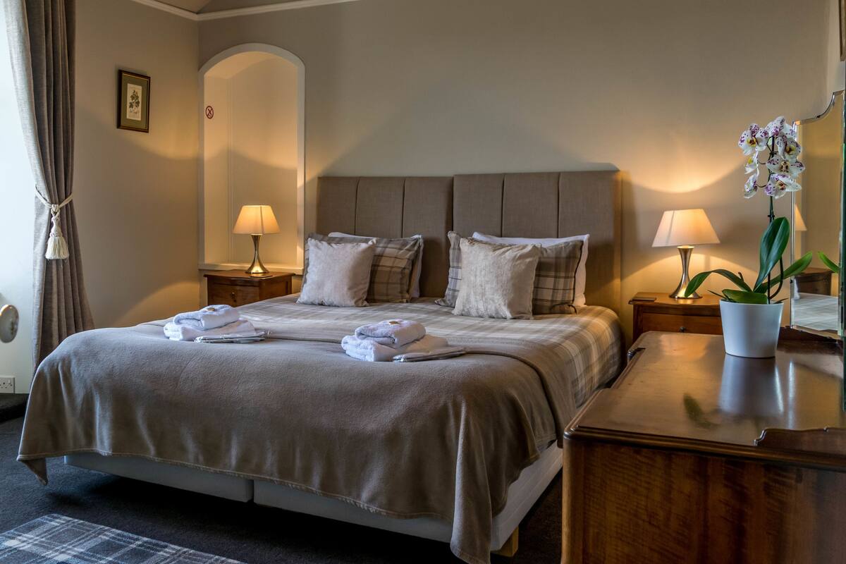 Uma cama bem arrumada com cabeceira cinza, duas mesas de cabeceira com abajures, uma orquídea em vaso e um quadro na parede.